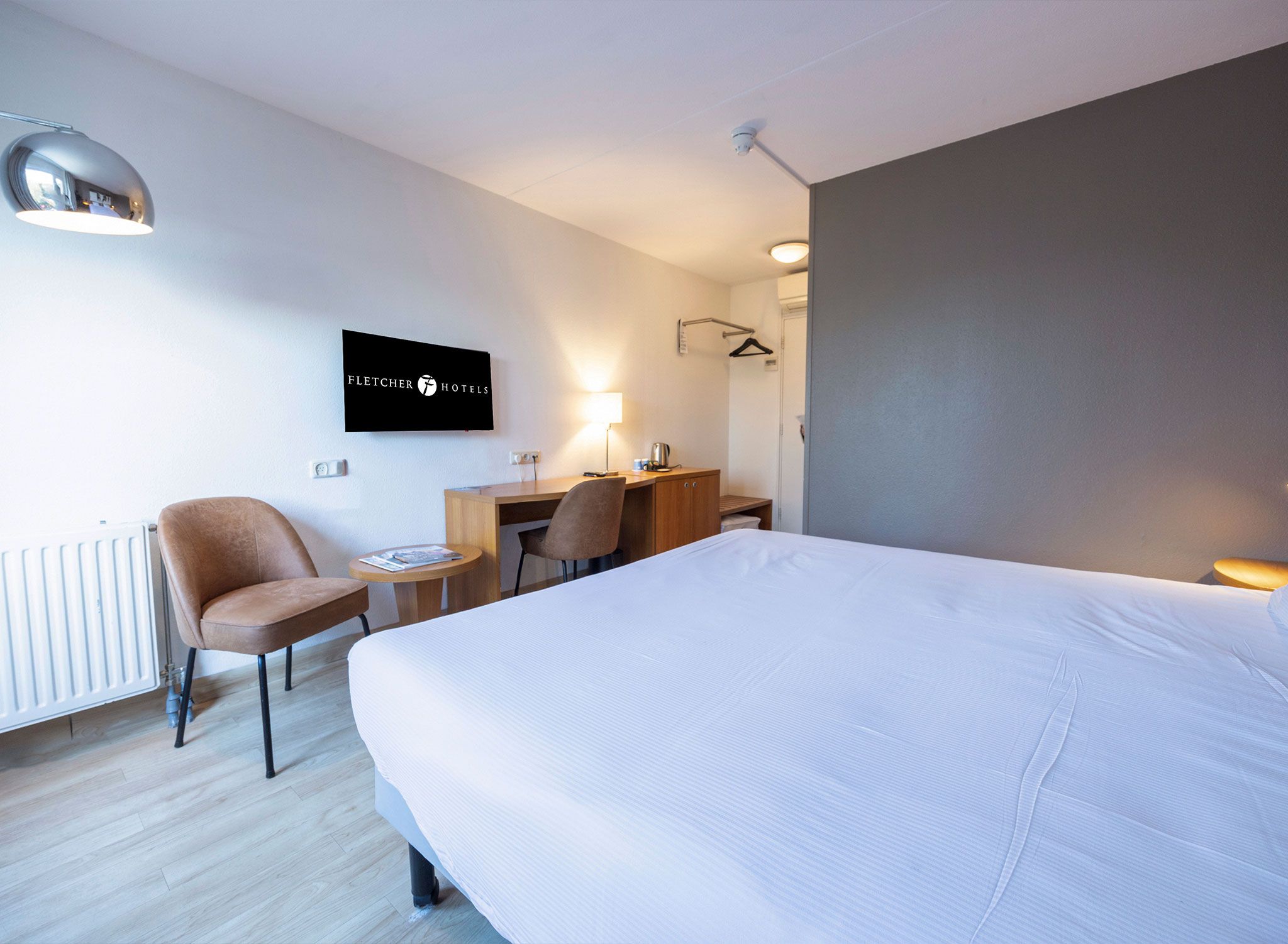 Quarto de hotel com cama, escrivaninha, cadeira, televisão e radiador.