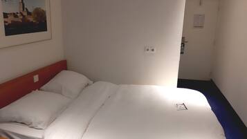 Quarto de hotel com uma cama, mesa de cabeceira, quadro na parede e porta com interruptor de luz.