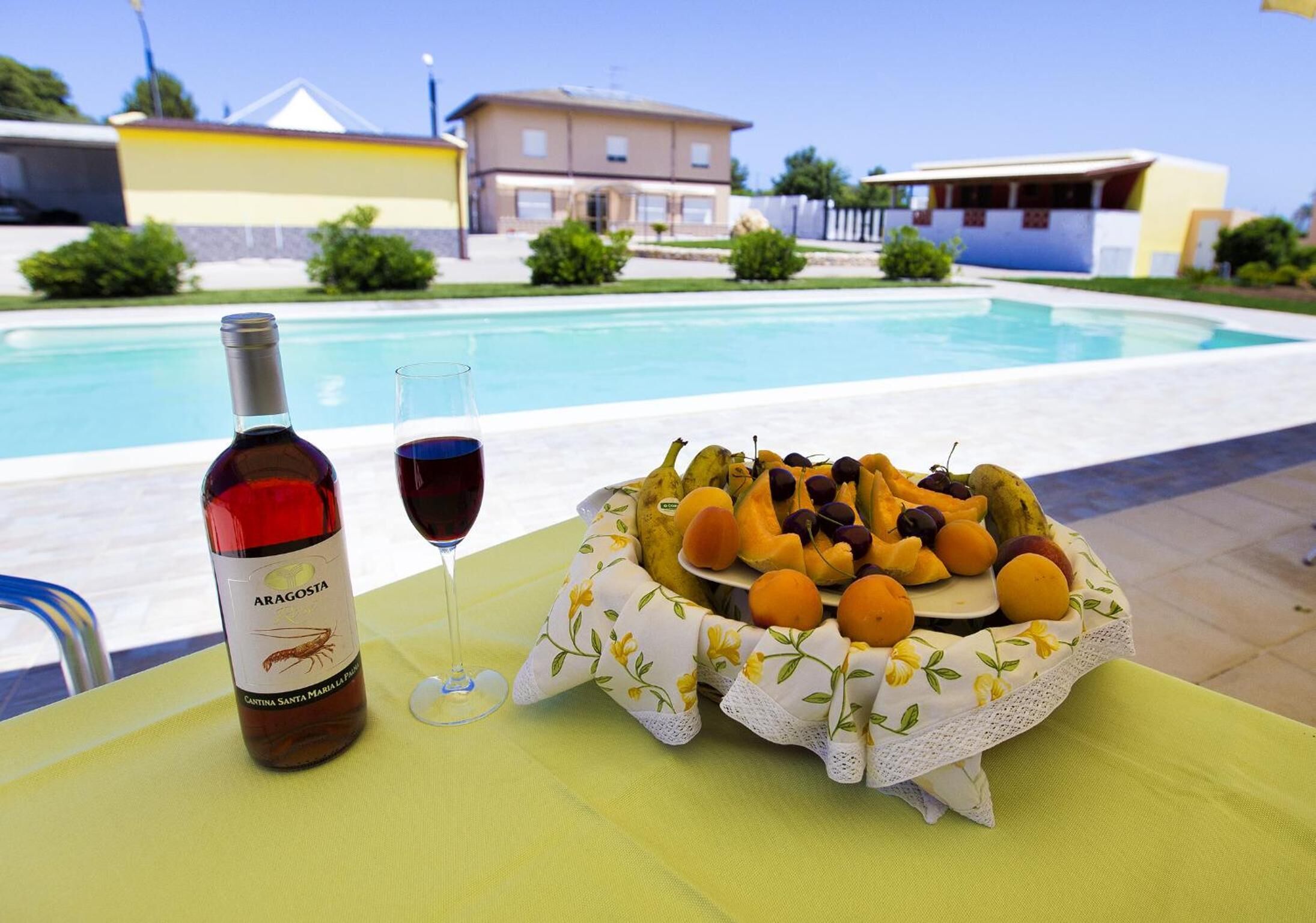 Uma mesa com uma garrafa de vinho Aragosta, um copo de vinho tinto e uma cesta de frutas junto a uma piscina.