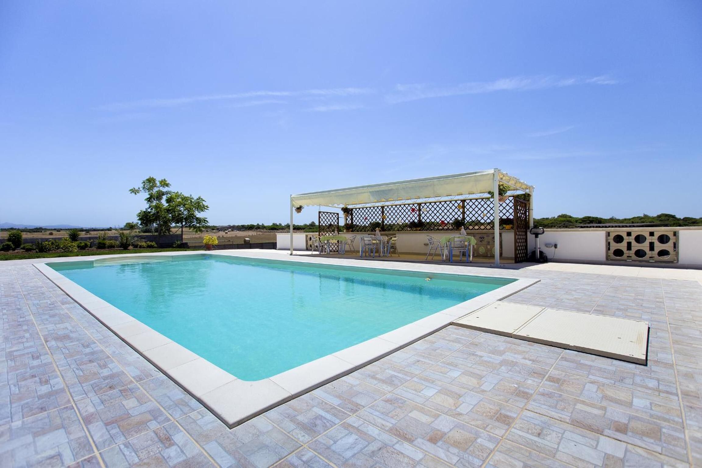 Piscina retangular com deck revestido, área de estar coberta com cadeiras e vista para a paisagem.