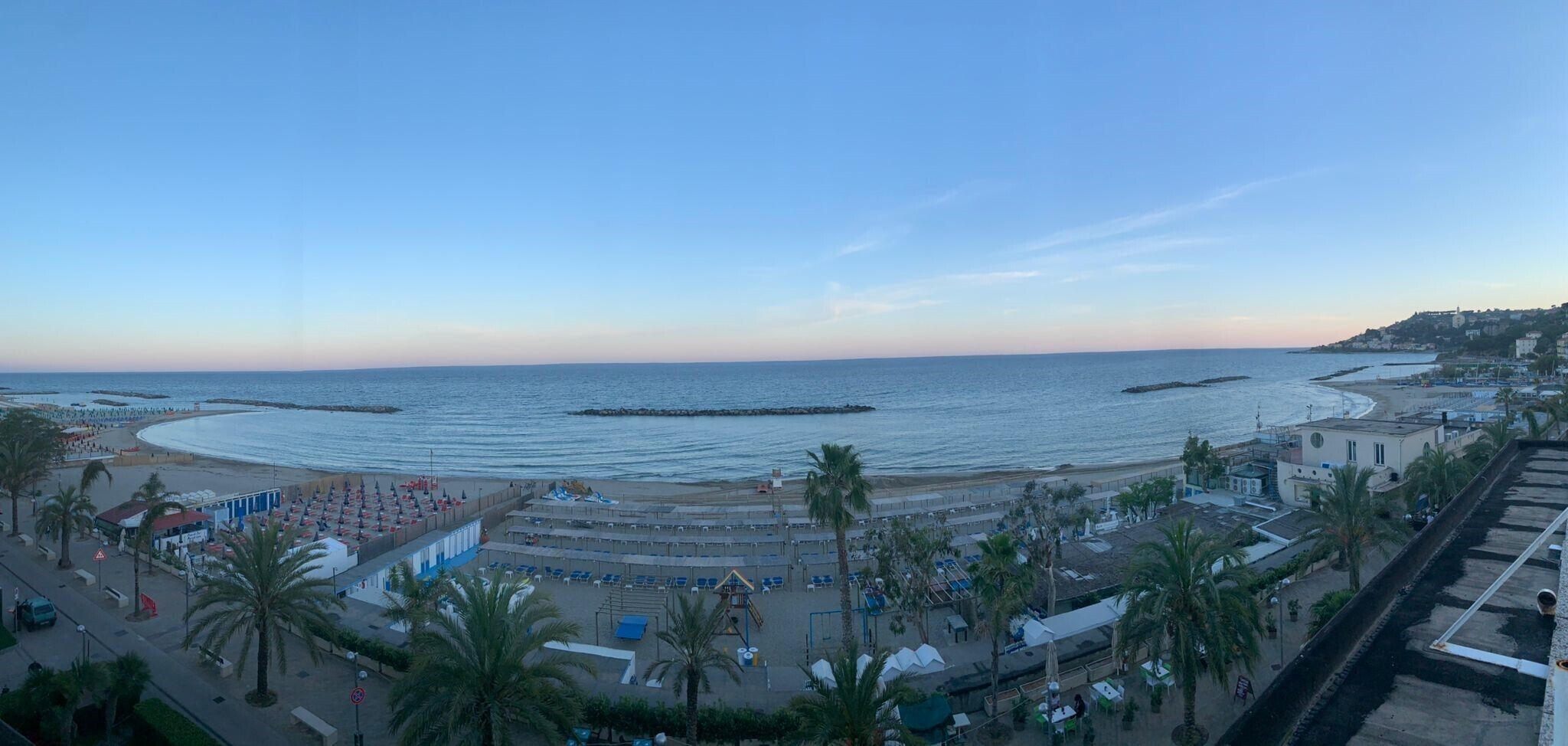Vista costeira com praia, palmeiras e calçadão com guarda-sóis.