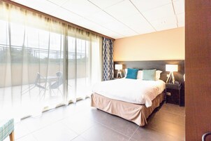 Quarto de hotel com uma cama grande, duas mesas de cabeceira com abajures, vista através de uma janela ampla e uma varanda com cadeiras.