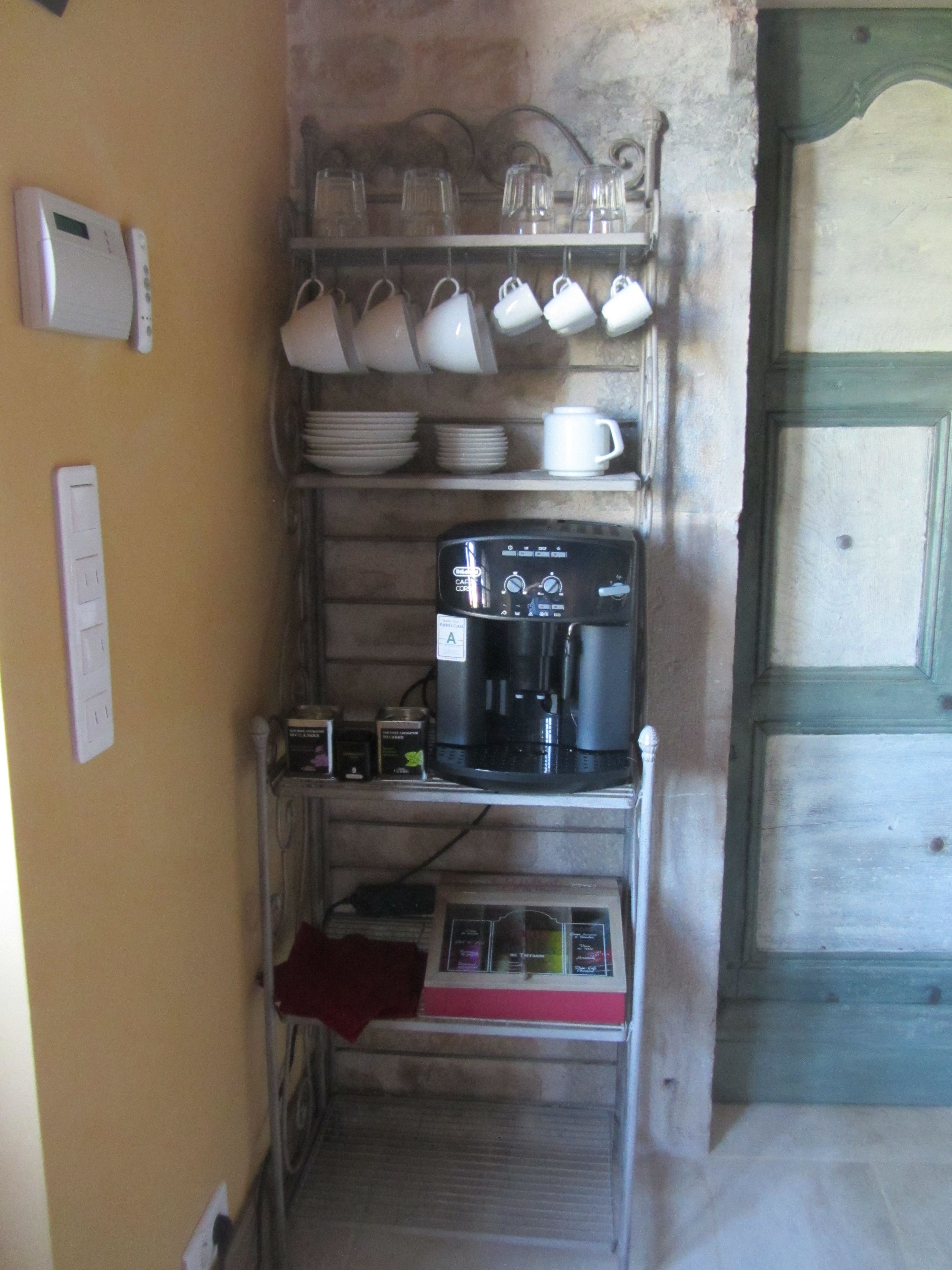 Uma cozinha compacta com cafeteira, prateleiras com xícaras e pratos, e uma geladeira pequena.
