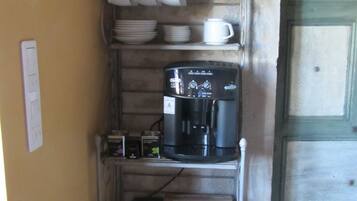 Uma cozinha compacta com cafeteira, prateleiras com xícaras e pratos, e uma geladeira pequena.