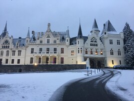 Um edifício grande e histórico com várias torres e torres pontiagudas, apresentando uma entrada imponente e uma entrada de carros coberta de neve.