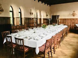 Uma longa mesa de jantar posta para uma refeição formal em um salão com painéis de madeira e janelas em arco.