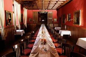 Mesa de jantar comprida com toalhas brancas e cadeiras, ladeada por paredes vermelhas e painéis de madeira.