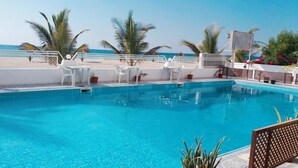 Uma piscina com água azul cristalina, rodeada por cadeiras e mesas brancas, palmeiras e vista para o oceano.