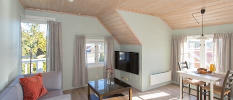 Uma sala de estar espaçosa, com teto alto, televisão de tela plana, mesa de centro de vidro, sofá com almofada vermelha, mesa de jantar com cadeiras e grandes janelas com cortinas.