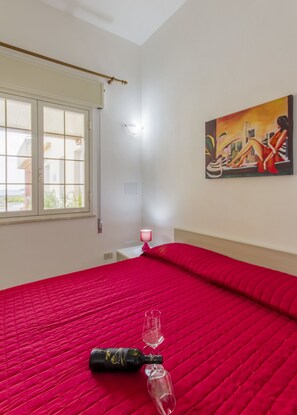 Quarto com uma cama grande, uma janela, um quadro e uma garrafa de vinho sobre a cama.