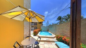 Uma área de piscina pequena ao ar livre com hidromassagem, cercada por um muro de tijolos e um edifício amarelo. Há uma mesa e cadeiras sob um toldo.