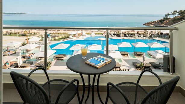 Varanda com vista para uma piscina e praia. Há cadeiras de praia e guarda-sóis.