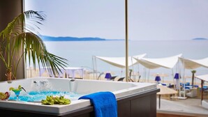Uma banheira de hidromassagem com uma toalha azul, um copo de martini e flores verdes numa varanda com vista para uma praia com guarda-sóis.