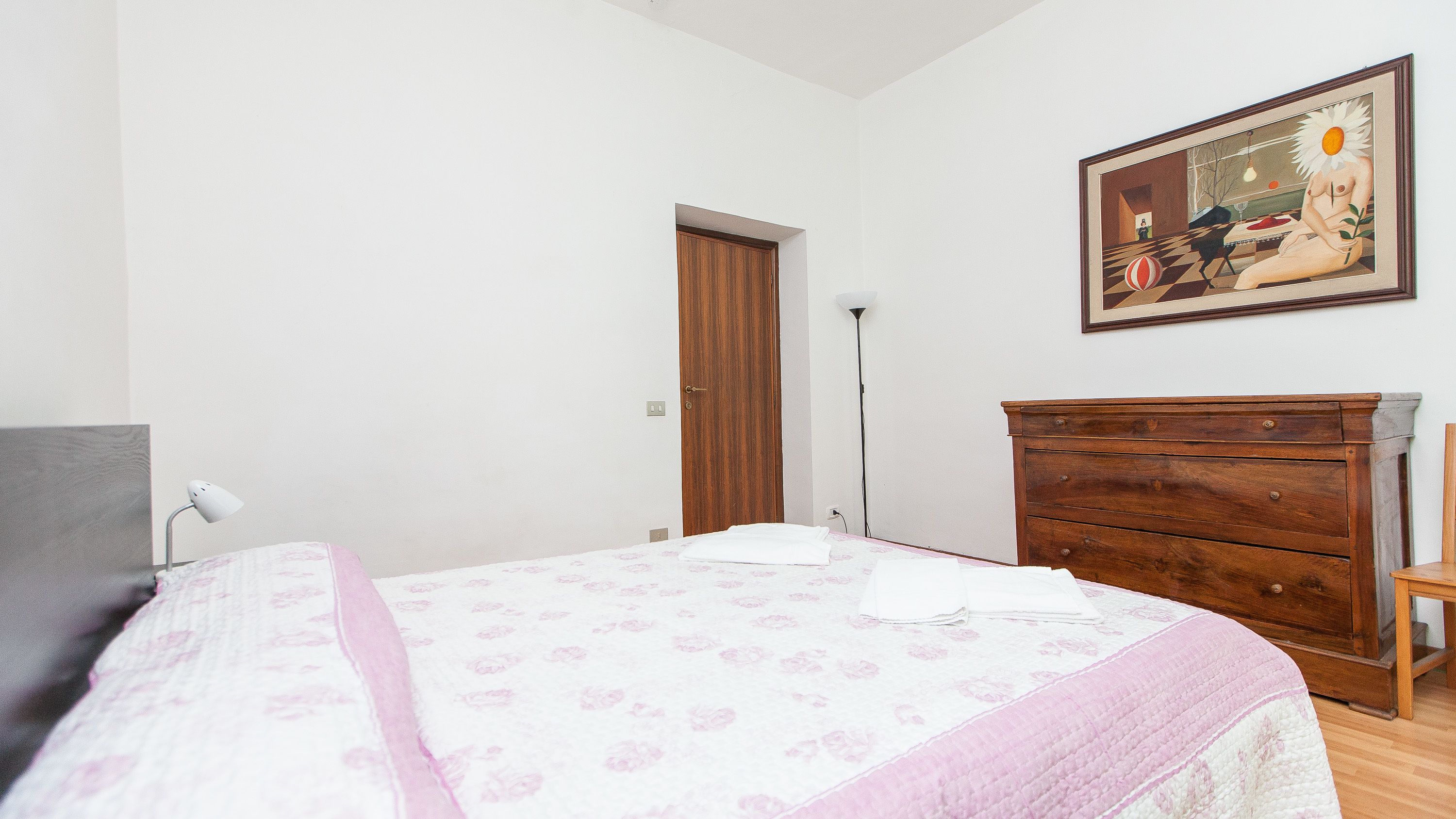 Quarto com cama, cómoda de madeira, porta e um quadro na parede.