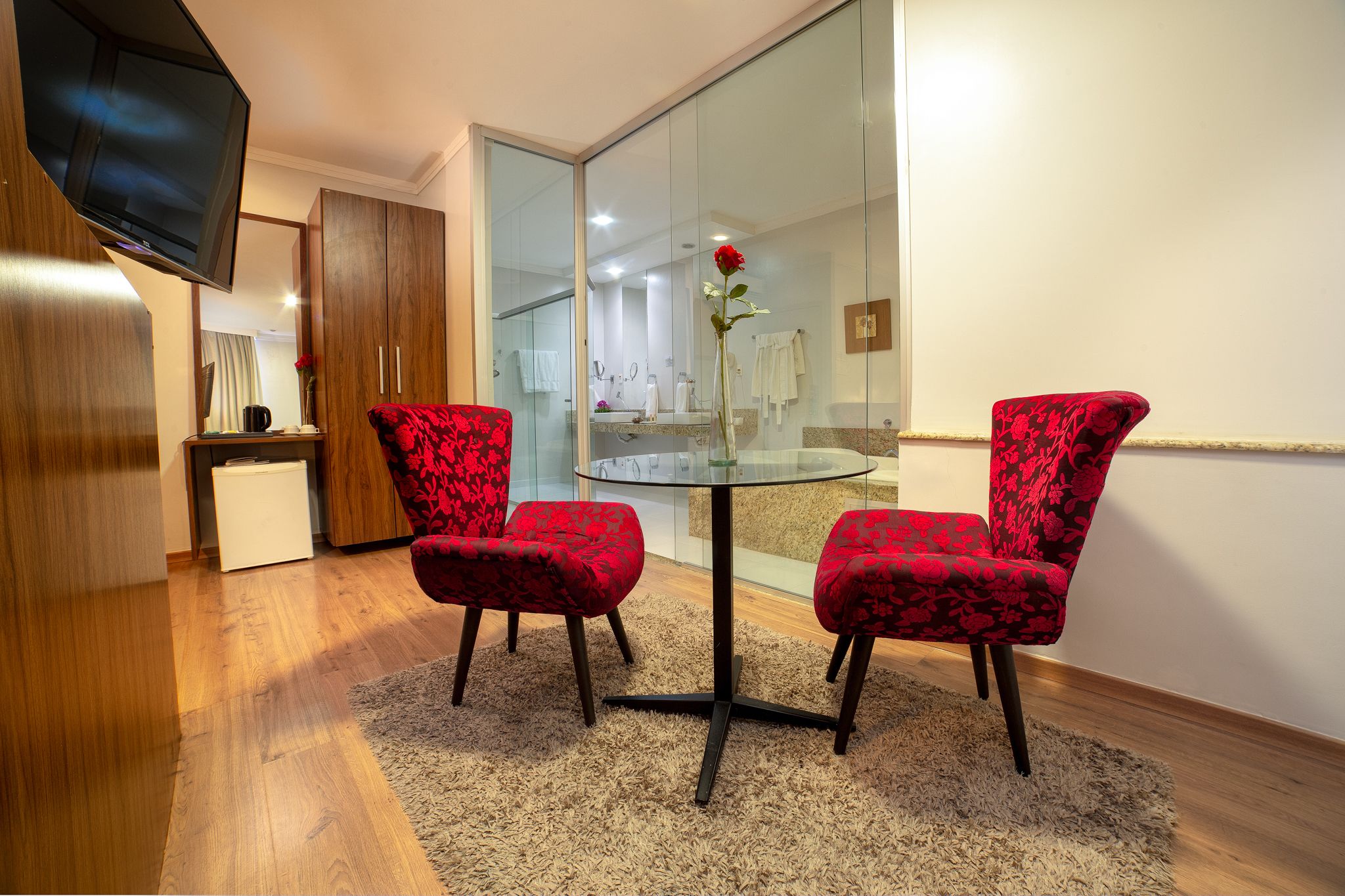 Quarto moderno com duas poltronas vermelhas ao redor de uma mesa de vidro, TV de tela plana na parede e vista para o banheiro através de um painel de vidro.