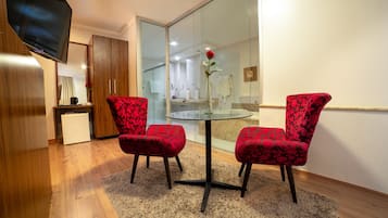 Quarto moderno com duas poltronas vermelhas ao redor de uma mesa de vidro, TV de tela plana na parede e vista para o banheiro através de um painel de vidro.