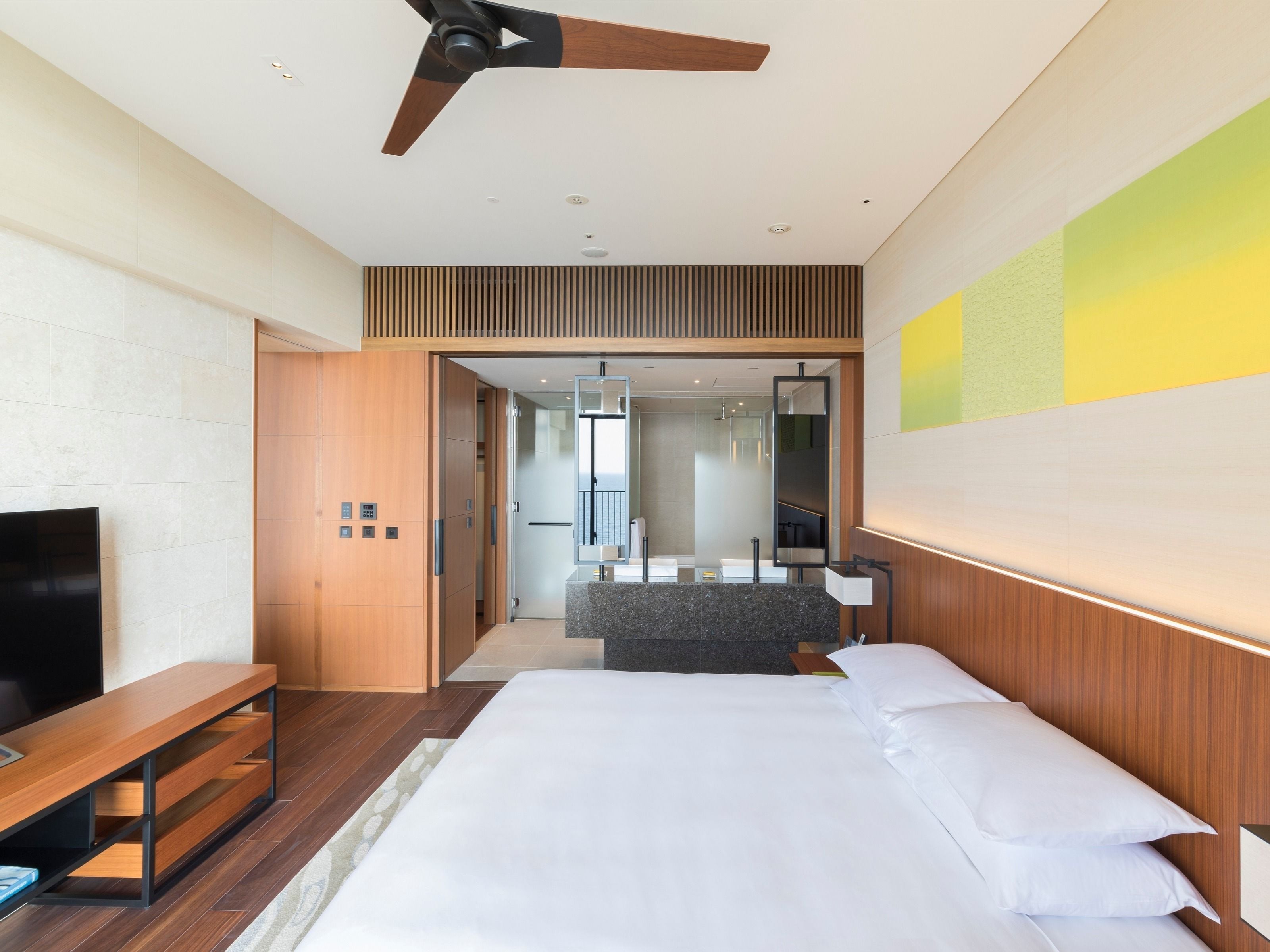 Quarto de hotel moderno com uma cama grande, uma televisão, um banco de madeira e um ventilador de teto.