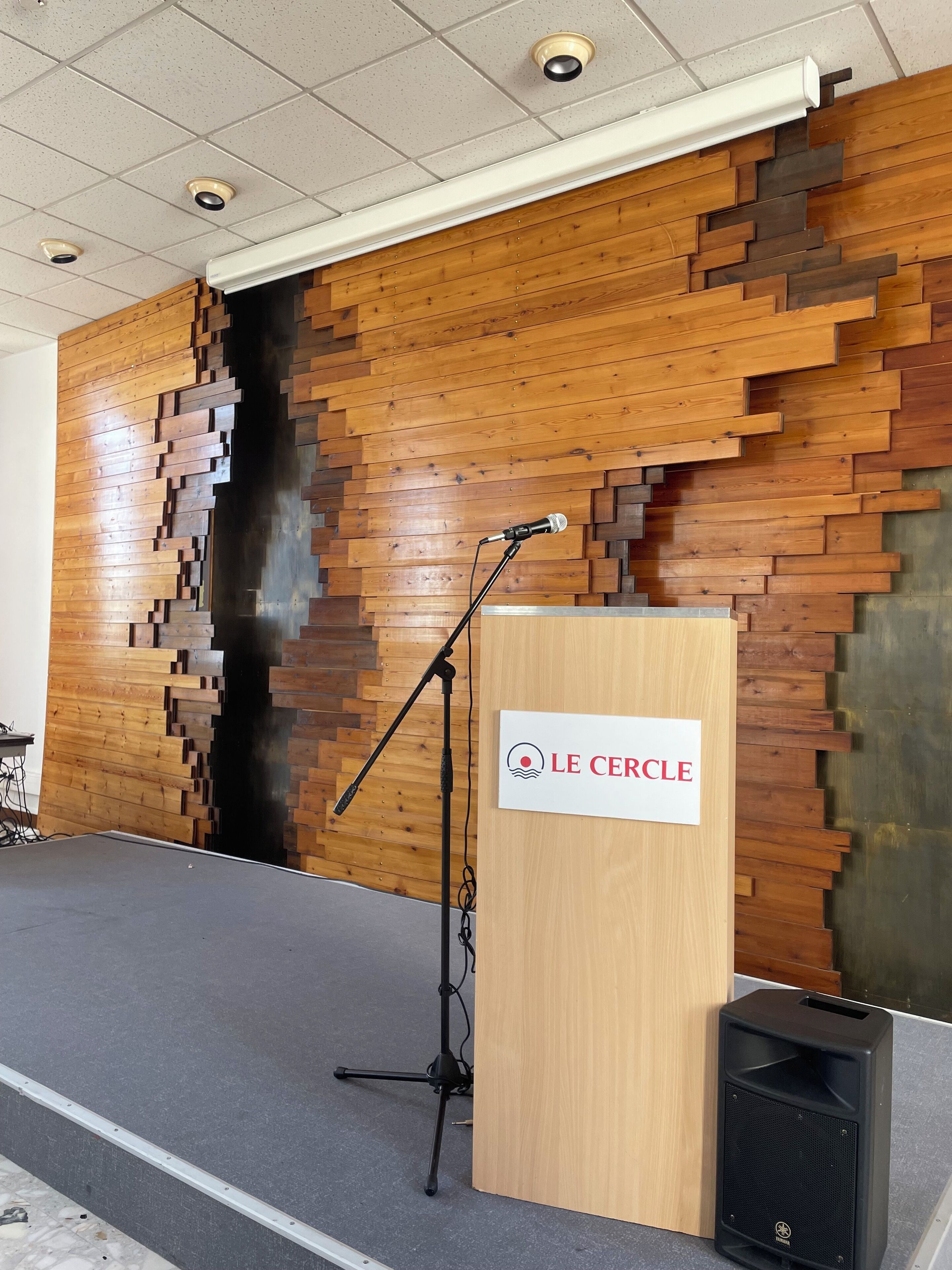 Uma sala de conferências com painel de parede em madeira, um pódio com a placa 'LE CERCLE' e um alto-falante no chão.