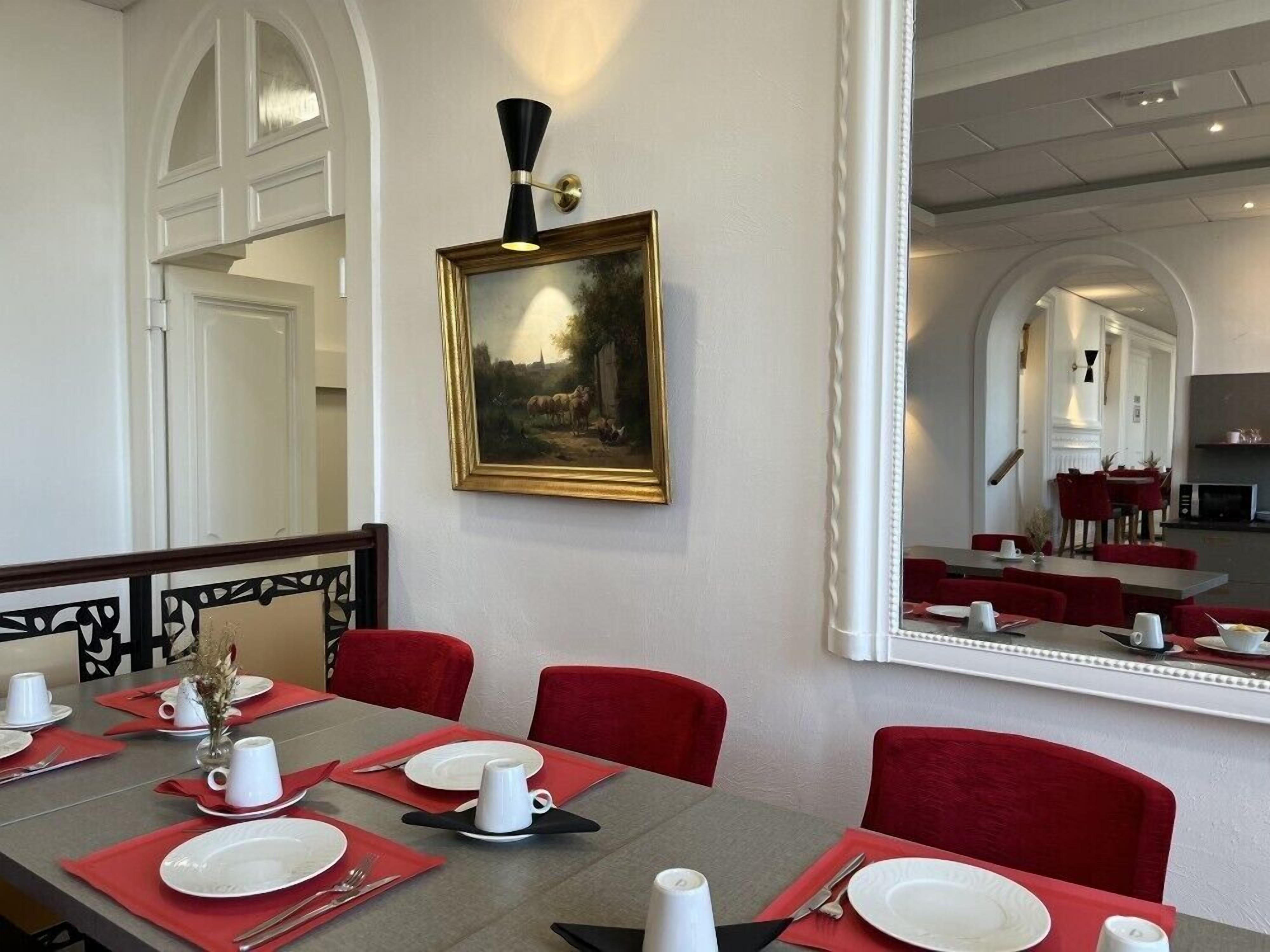 Uma área de refeições com uma mesa posta para quatro pessoas, cadeiras vermelhas, um quadro na parede e um espelho grande que reflete o ambiente.