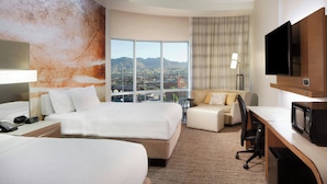 Quarto de hotel com duas camas, uma mesa de trabalho com televisão, luminária de mesa e vista para a cidade.