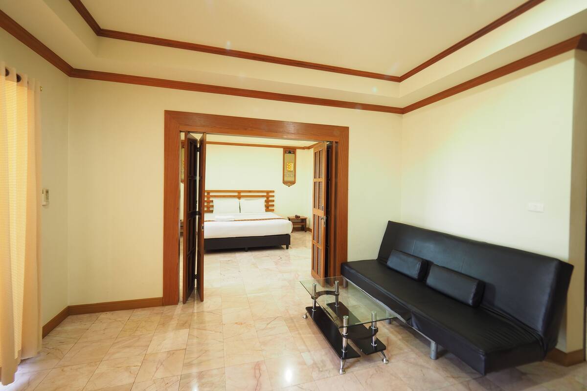 Quarto de hotel com cama, sofá de couro preto, mesa de centro de vidro e porta de madeira que dá para outro quarto.