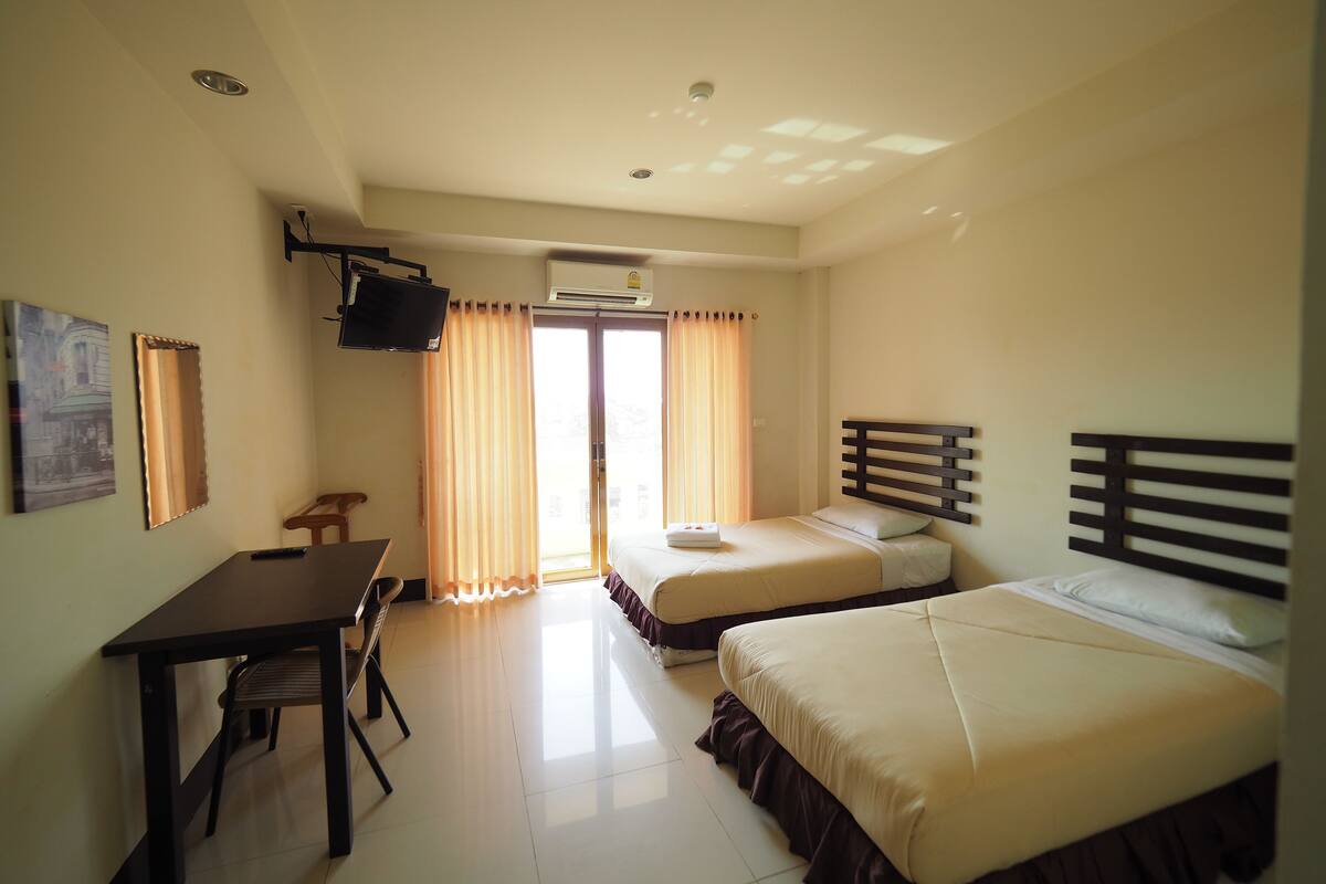 Quarto de hotel com duas camas, uma escrivaninha, uma cadeira, ar condicionado e uma televisão na parede.