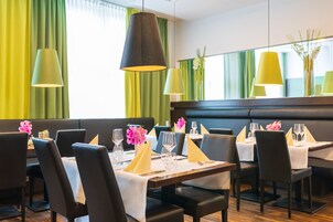 Restaurante com mesas postas, cortinas verdes e luminárias pendentes.