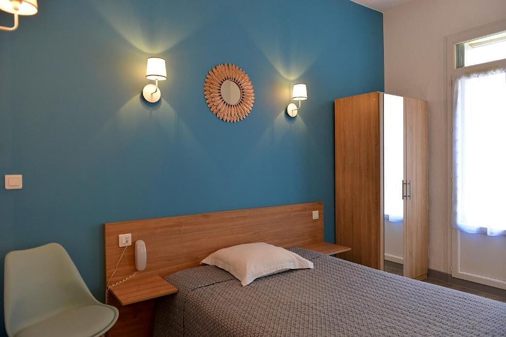 Quarto com parede azul, cabeceira de madeira, cama com manta estampada, mesa de cabeceira com abajur, espelho e guarda-roupa.