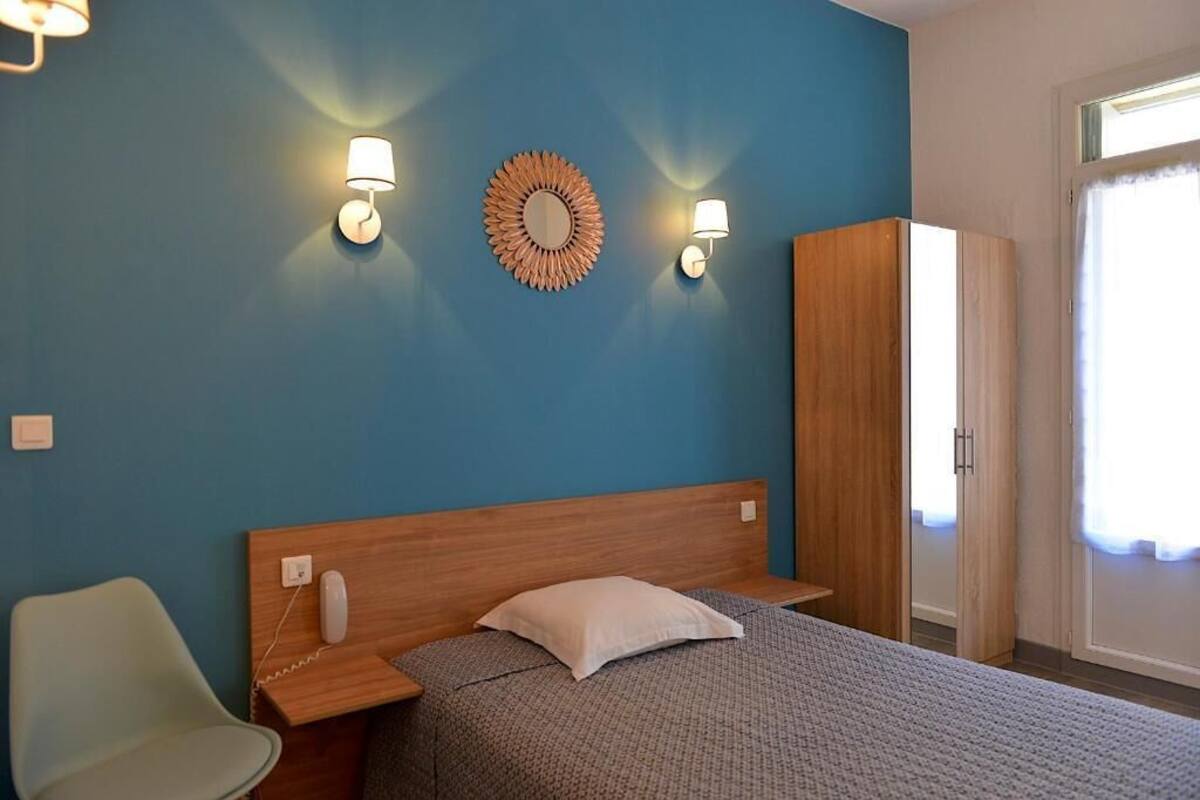 Quarto com parede azul, cabeceira de madeira, cama com manta estampada, mesa de cabeceira com abajur, espelho e guarda-roupa.
