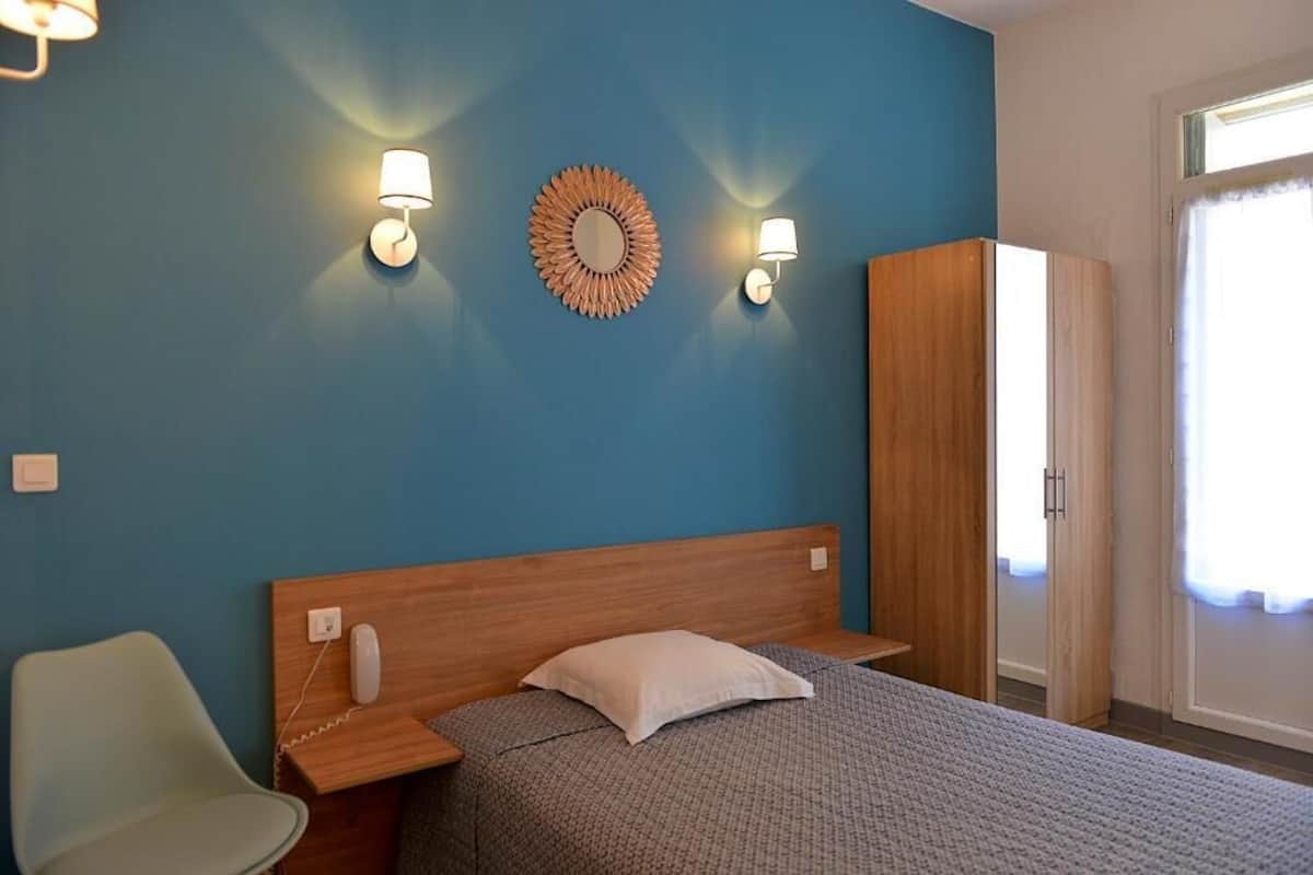 Quarto com parede azul, cabeceira de madeira, cama com manta estampada, mesa de cabeceira com abajur, espelho e guarda-roupa.