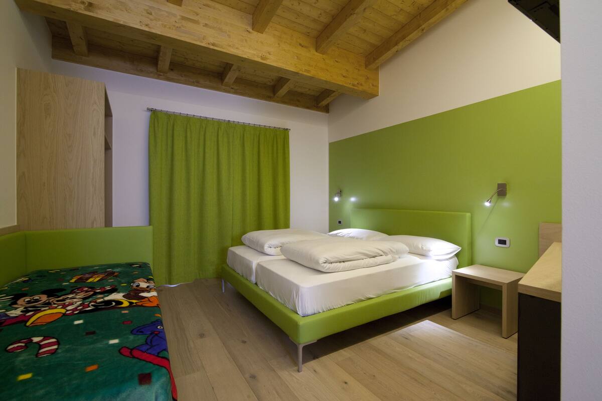Quarto com cama, mesa de cabeceira, cortina verde e teto de madeira.