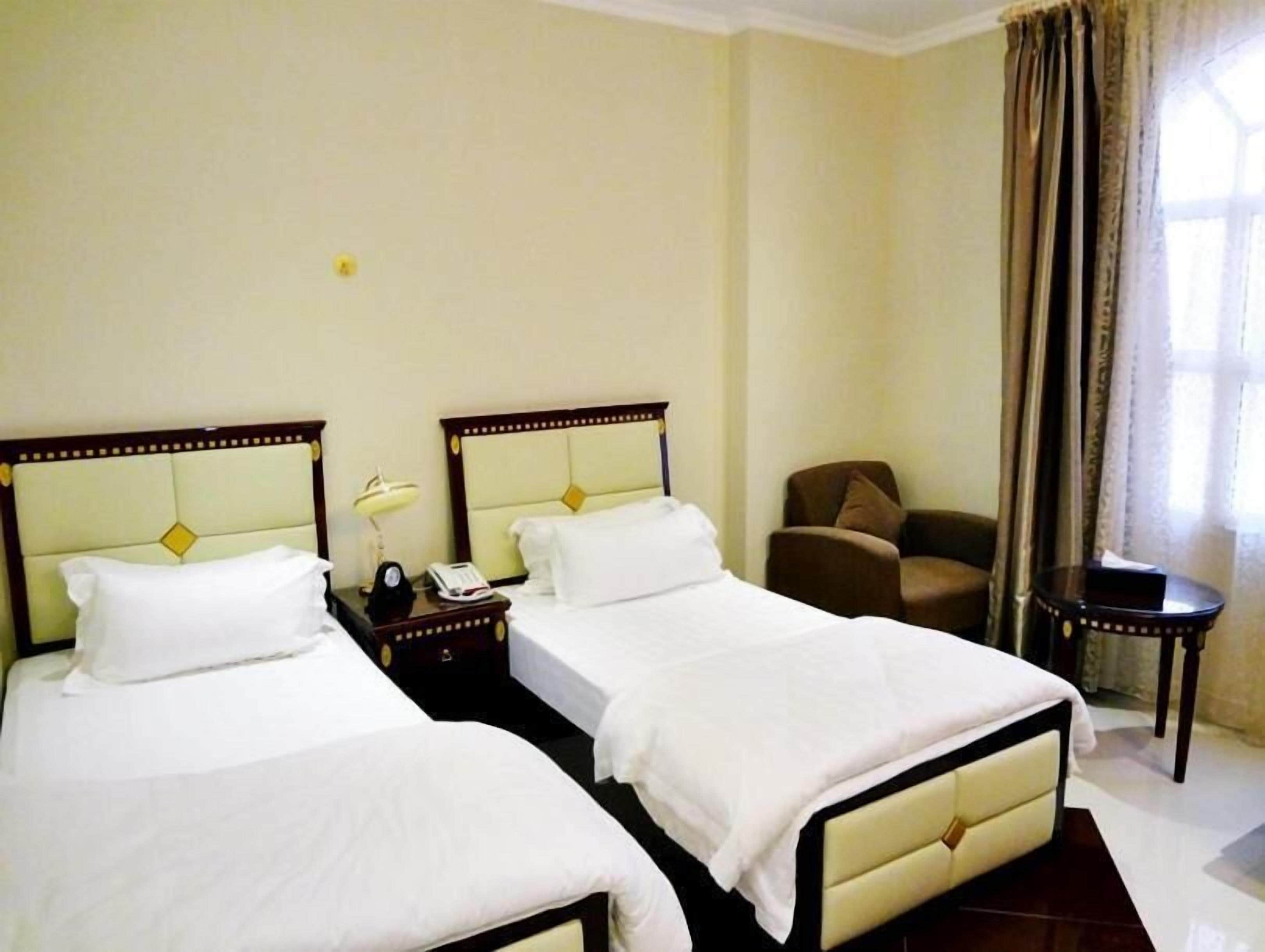 Quarto de hotel com duas camas, uma cadeira, uma mesinha e um criado-mudo com abajur e telefone.