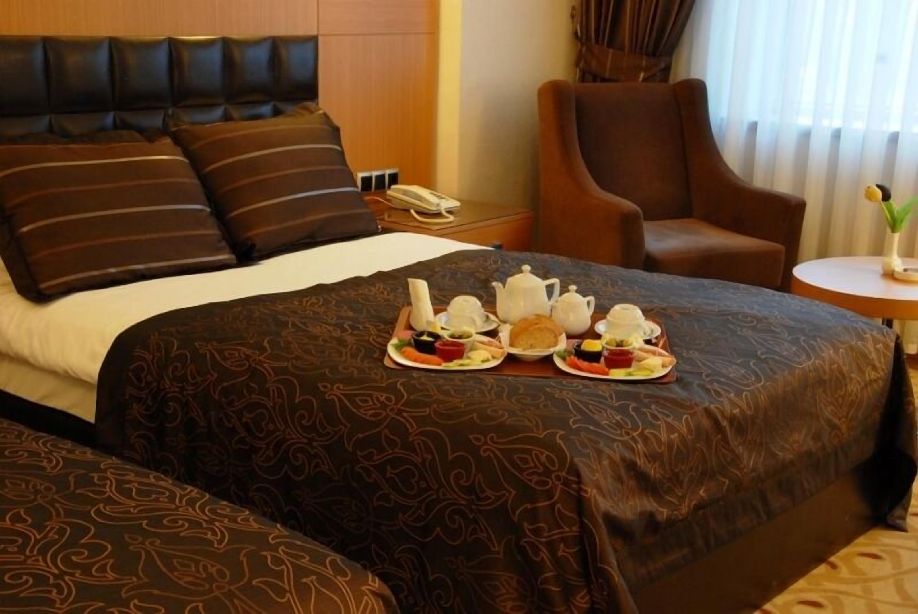 Quarto de hotel com uma cama grande, uma mesa de cabeceira com telefone, uma poltrona marrom e uma pequena mesa com um vaso e uma planta.