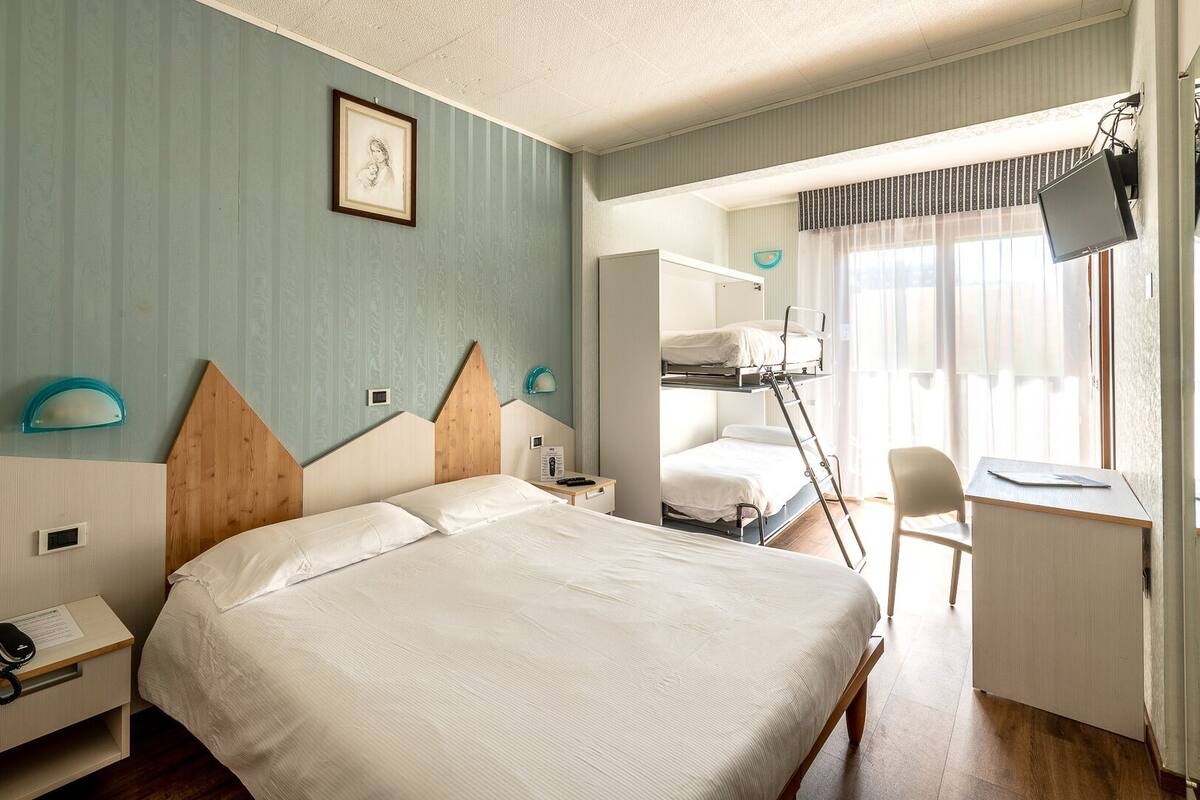 Quarto de hotel com cama, mesa de cabeceira, escrivaninha, cadeira, beliches e uma janela com cortinas.