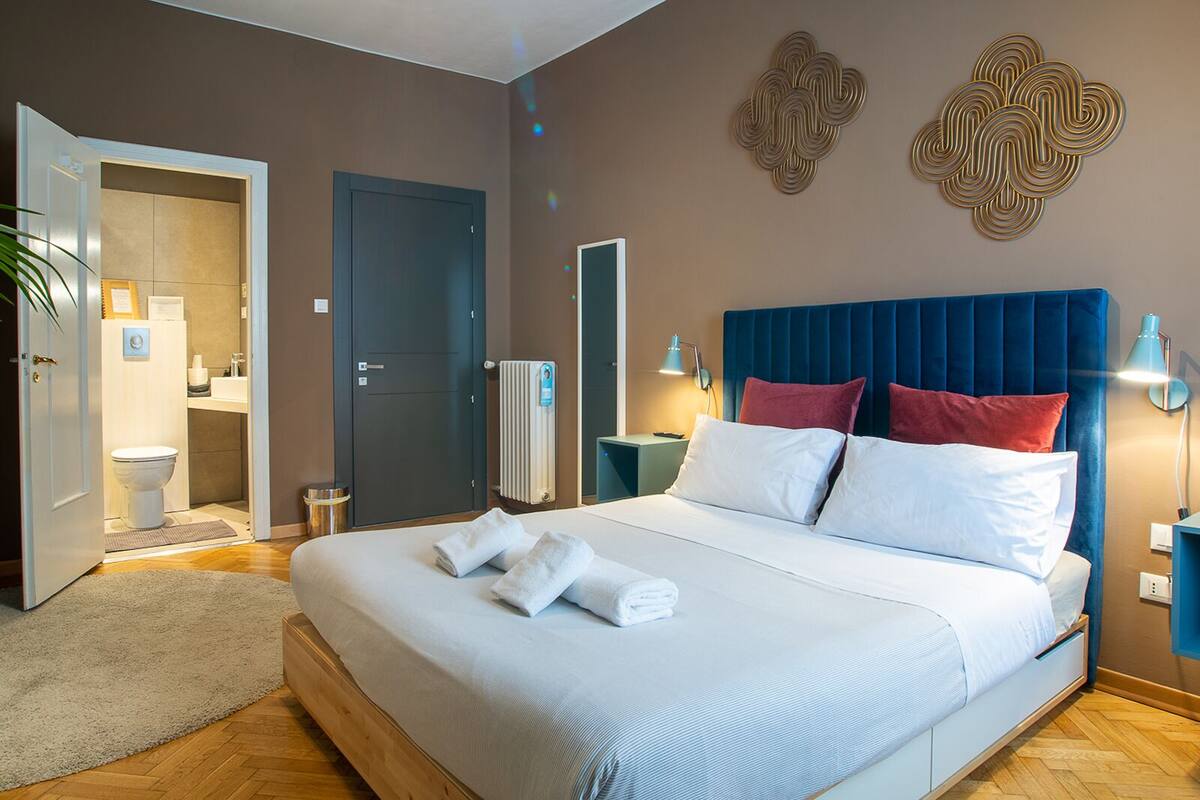 Quarto de hotel com uma cama grande, uma mesa de cabeceira, um radiador, uma luminária na parede e uma porta que dá para o banheiro.