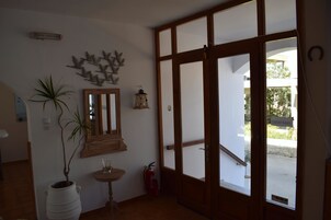 Quarto com porta de madeira grande, uma mesa pequena com um copo, uma planta em vaso e um espelho fixado na parede.