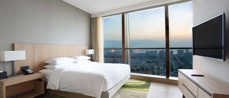 Quarto de hotel moderno com uma cama grande, uma mesinha de cabeceira com luminária, TV fixada na parede e vista para a cidade através de grandes janelas.
