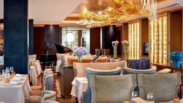 Restaurante com mesas postas para refeições, cadeiras, um piano de cauda e um lustre decorativo.