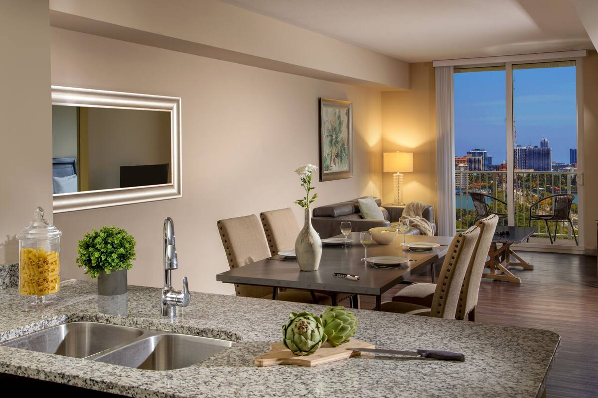 Quarto de hotel moderno com área de refeições, cozinha com bancadas de granito, pia e uma grande janela com vista para a cidade.