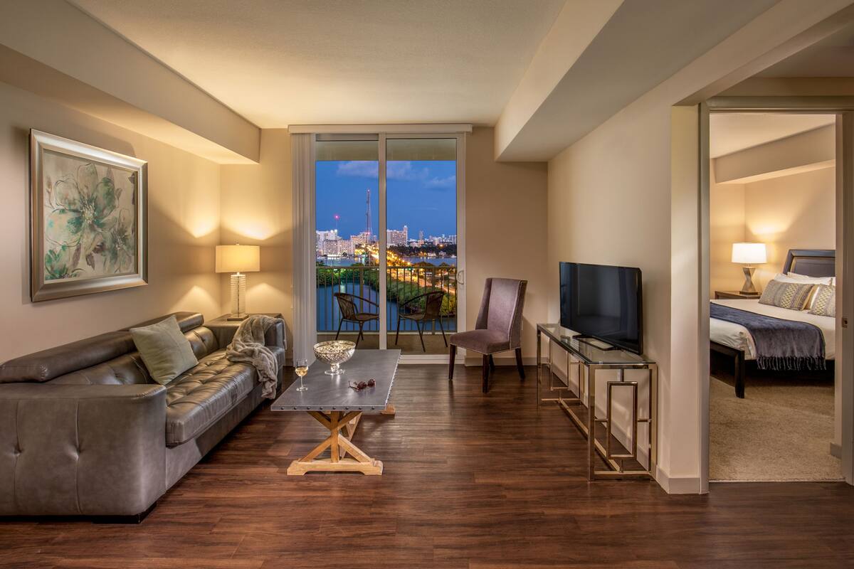 Sala de estar moderna com sofá, mesa de vidro, televisão e varanda com vista para a cidade.