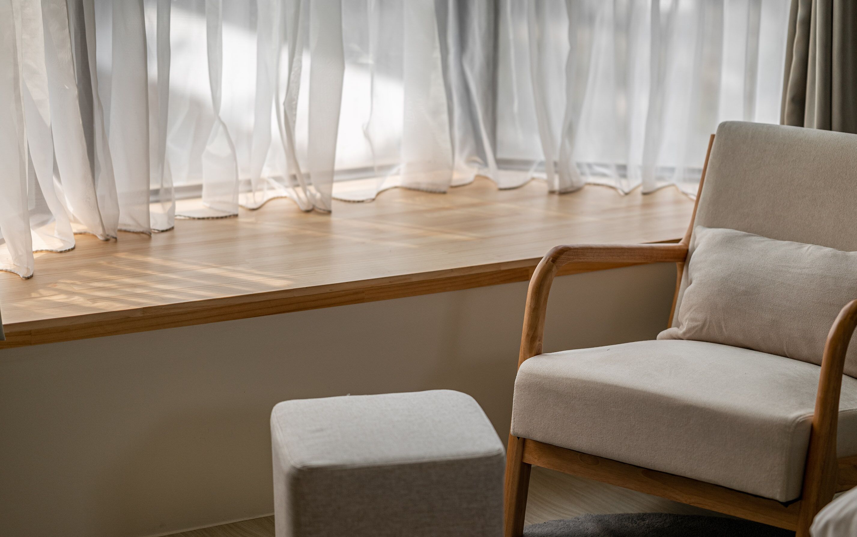 Uma sala de estar moderna com um banco de madeira, uma poltrona estofada e cortinas leves.
