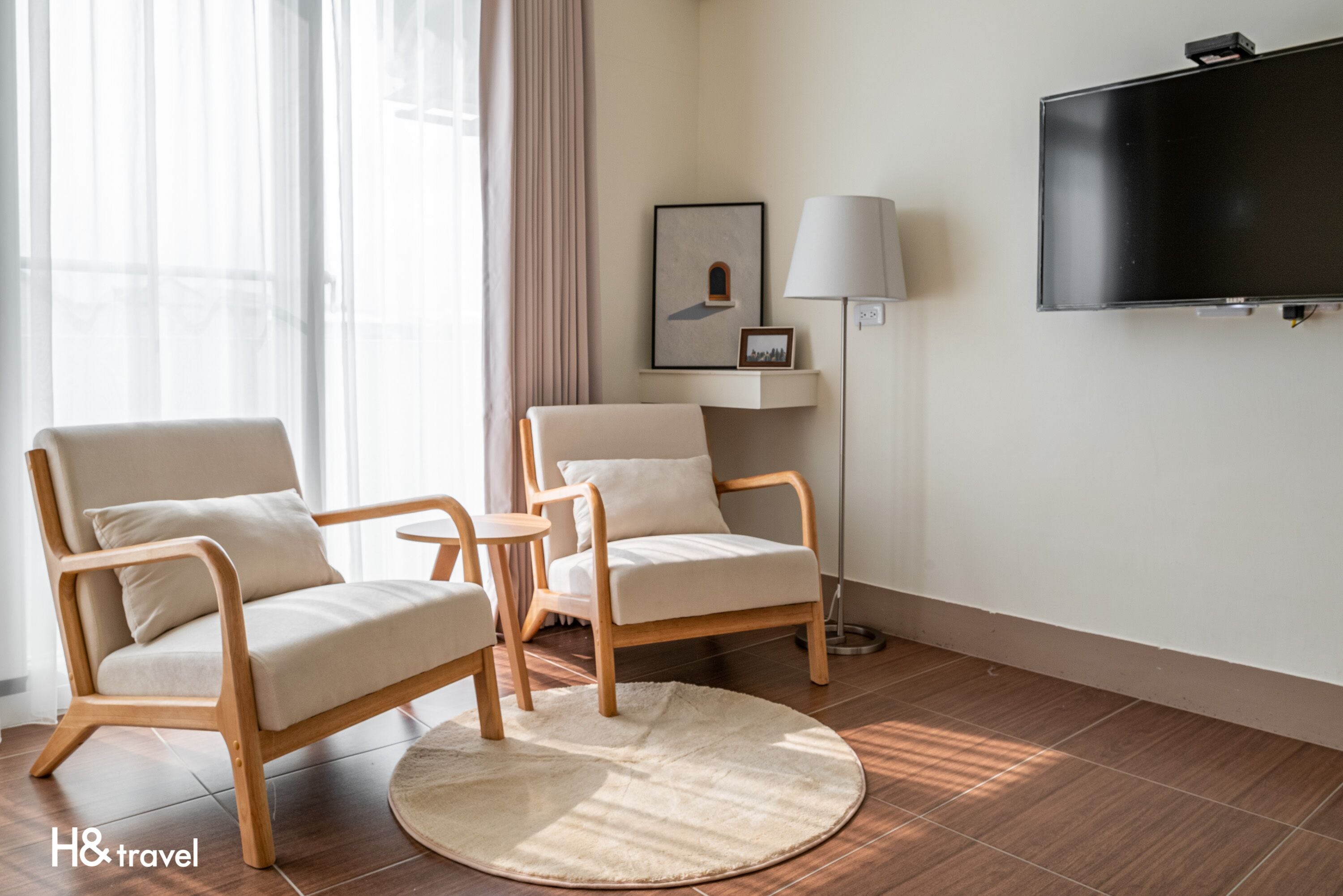 Quarto de hotel com duas cadeiras de madeira, um tapete redondo, uma televisão, um abajur e um quadro na parede.