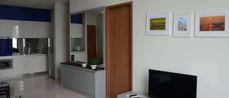 Uma sala de estar moderna com uma televisão de tela plana, uma mesa de centro com um vaso e um sofá. Há uma área de cozinha com armários e uma geladeira ao fundo.