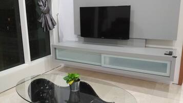 Uma sala de estar moderna com uma mesa de centro de vidro, uma televisão de tela plana presa a uma parede branca e uma porta de vidro deslizante com cortinas.