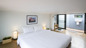 Quarto moderno com uma cama grande, luminárias de cabeceira, um quadro emoldurado, uma mesinha de madeira com uma planta e vista para um banheiro através de uma porta de vidro.
