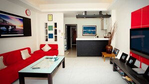 Uma sala de estar moderna com um sofá vermelho, uma mesa de centro de vidro e uma TV de tela plana. Há uma área de cozinha com bancada preta e um armário vermelho.