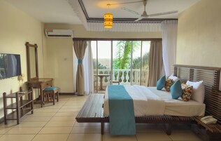 Quarto de hotel com cama, escrivaninha, cadeira, televisão e varanda com vista para palmeiras.