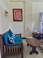 Quarto com sofá de madeira, uma pequena mesa de madeira, um quadro abstrato emoldurado, uma luminária na parede e um pequeno objeto decorativo em uma mesinha de cabeceira de madeira.