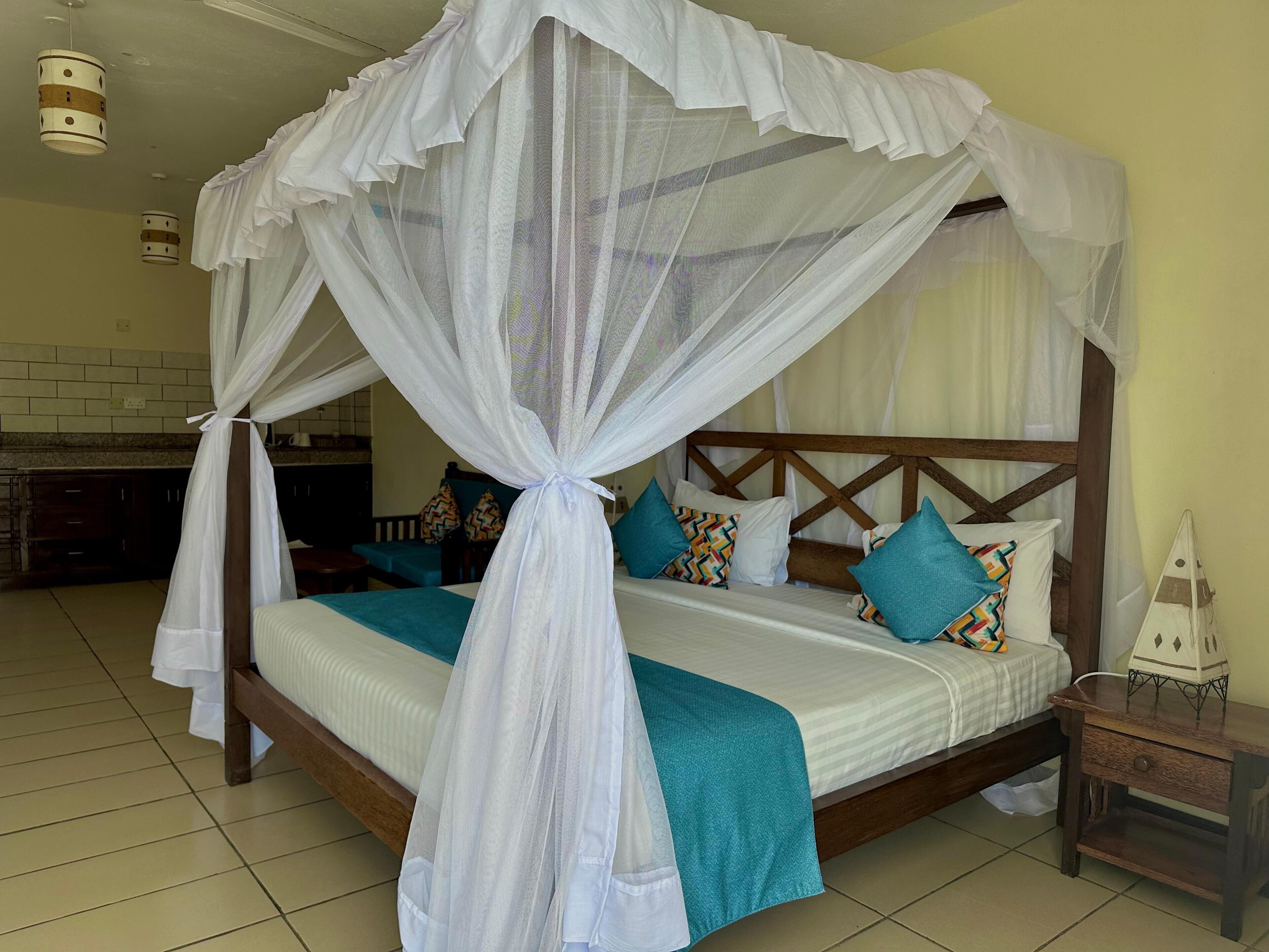 Uma cama com dossel e cortinas, cabeceira de madeira, manta azul-esverdeada e almofadas estampadas.