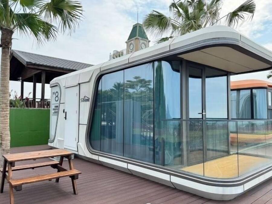 Estrutura moderna em vidro curvo com um banco e uma mesa, provavelmente uma área de descanso ou espera, com vista para palmeiras e um edifício ao fundo.